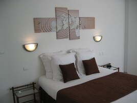 Cama bem arrumada com roupa de cama branca e castanha, duas luminárias fixadas na parede e três obras de arte na parede acima da cama.