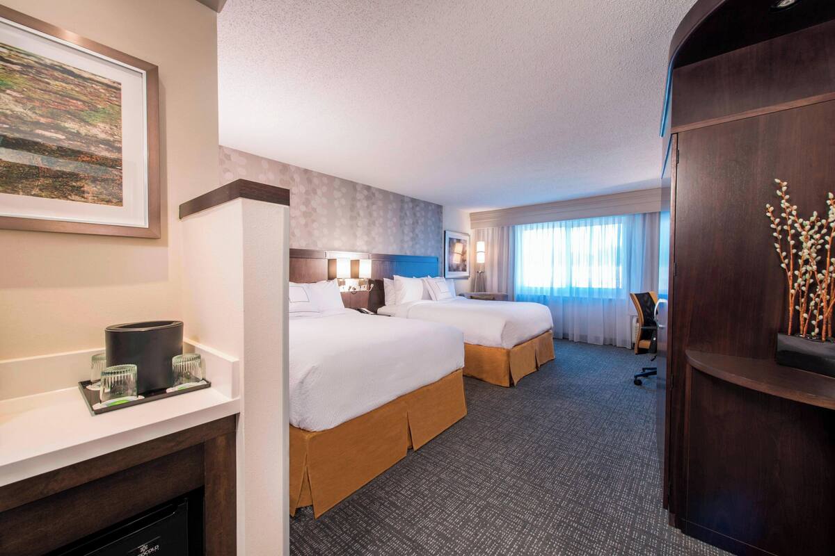 Quarto de hotel com duas camas, uma escrivaninha, uma cadeira, lareira e uma grande janela com cortinas.