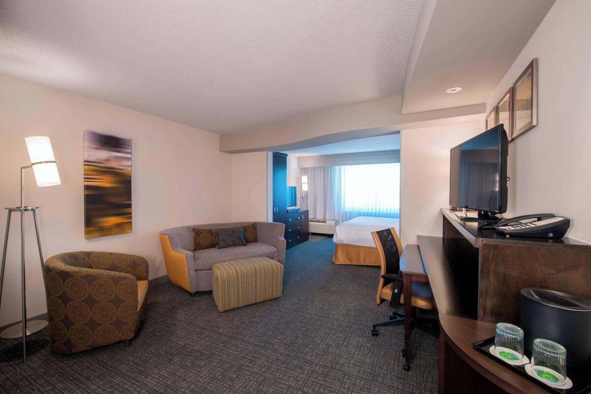 Quarto de hotel com sofá, poltrona, escrivaninha e televisão.