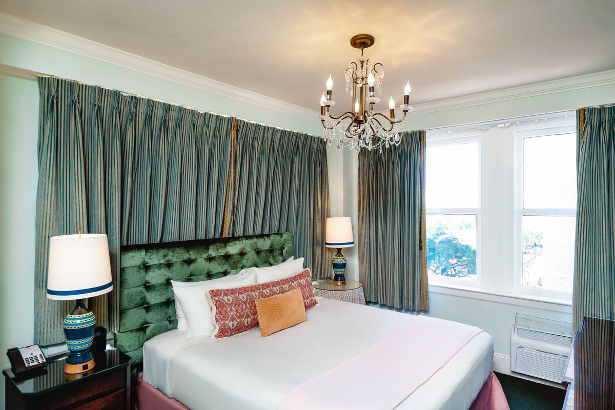 Quarto de hotel com uma cama grande, duas mesas de cabeceira com abajures, um lustre e uma janela com cortinas.