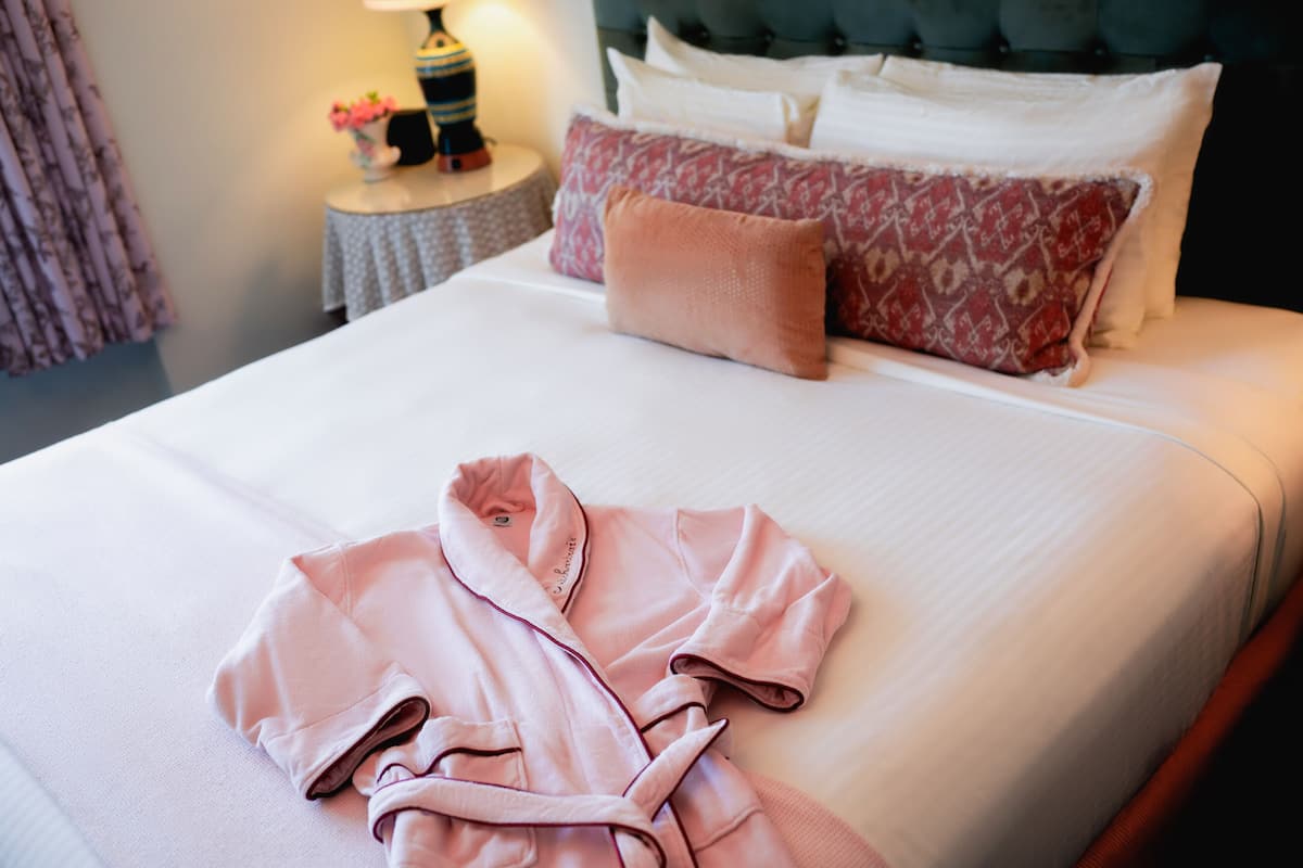 Cama bem arrumada com um roupão rosa, almofada estampada e mesa de cabeceira com abajur e flores.