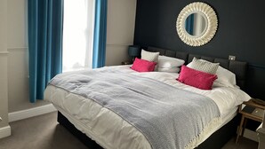 Quarto com cama grande, espelho redondo e janela com cortinas azuis.