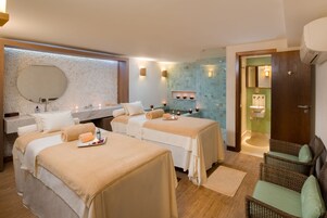 Sala de spa com duas camas, uma mesa de massagem, um espaço de estar pequeno e um banheiro com pia e espelho.