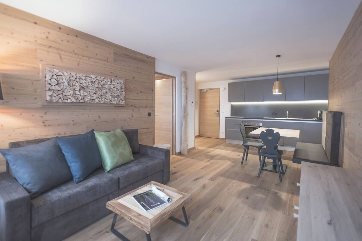 Um interior moderno com um sofá cinza, uma mesa de centro de madeira, uma área de jantar com uma cadeira e uma cozinha com armários cinzas e uma luminária pendente.