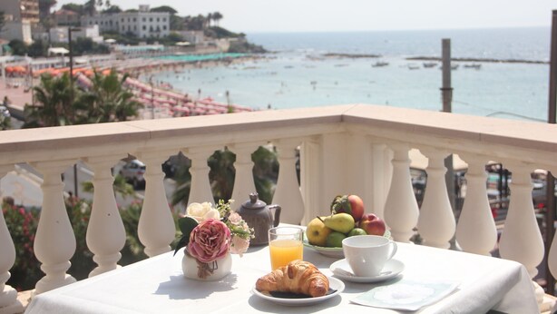 Mesa na varanda com itens de café da manhã, vista para a praia e paisagem urbana ao fundo.