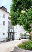Um edifício branco com entrada central, várias janelas e uma árvore em primeiro plano.