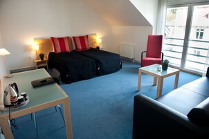 Quarto de hotel com cama, uma cadeira vermelha, uma pequena mesa com uma planta e uma escrivaninha com abajur e chaleira.