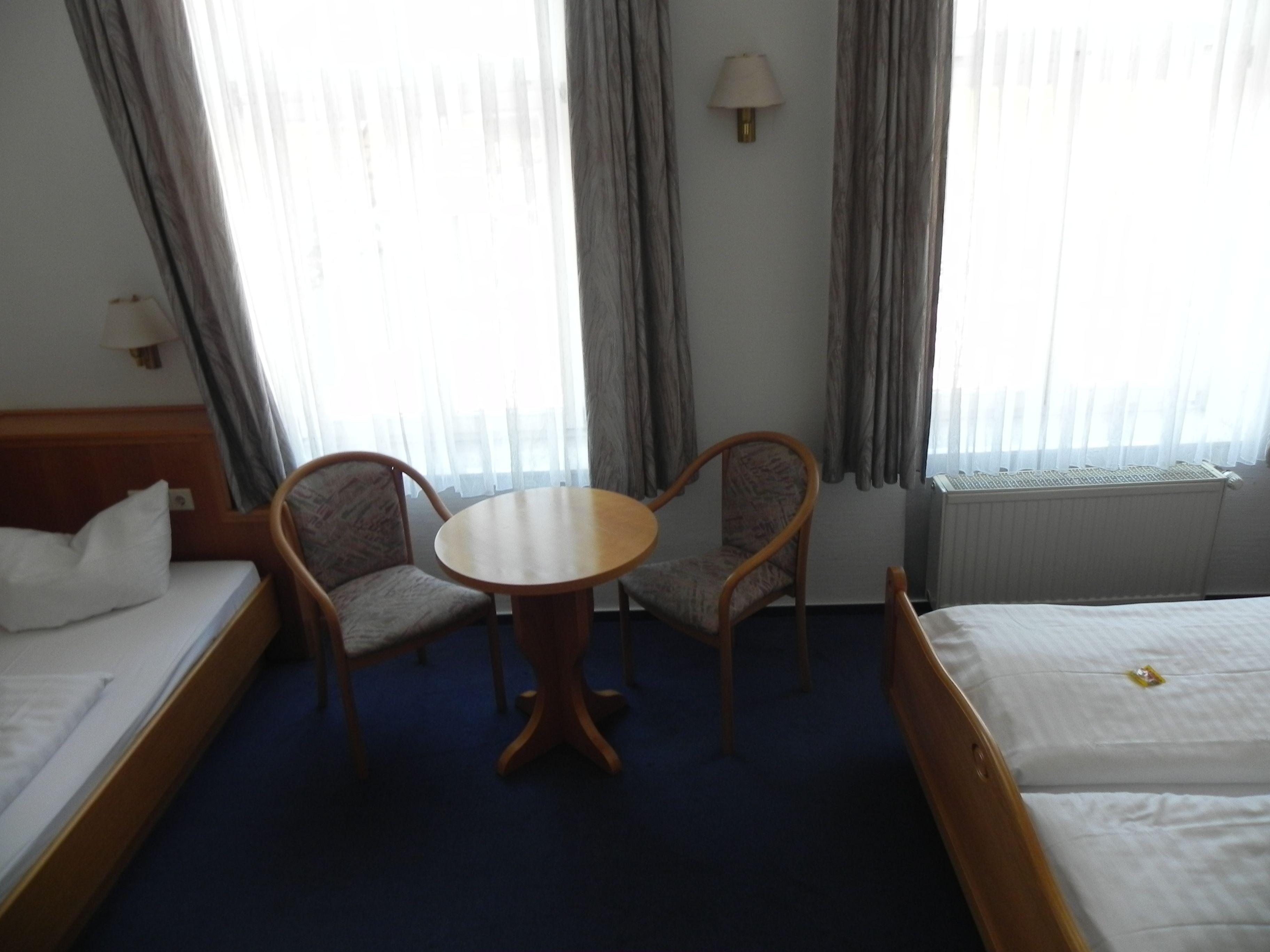 Quarto de hotel com duas camas de solteiro, uma mesa redonda, duas cadeiras, uma luminária e uma janela com cortinas.