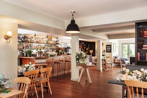 Uma área de bar com banquetas, balcão e diversas garrafas. Cadeiras de madeira e uma mesa com flores.