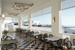 Um restaurante com piso xadrez, cadeiras e mesas de metal preto, amplas janelas com vista para a cidade e uma escada que leva a um andar superior.