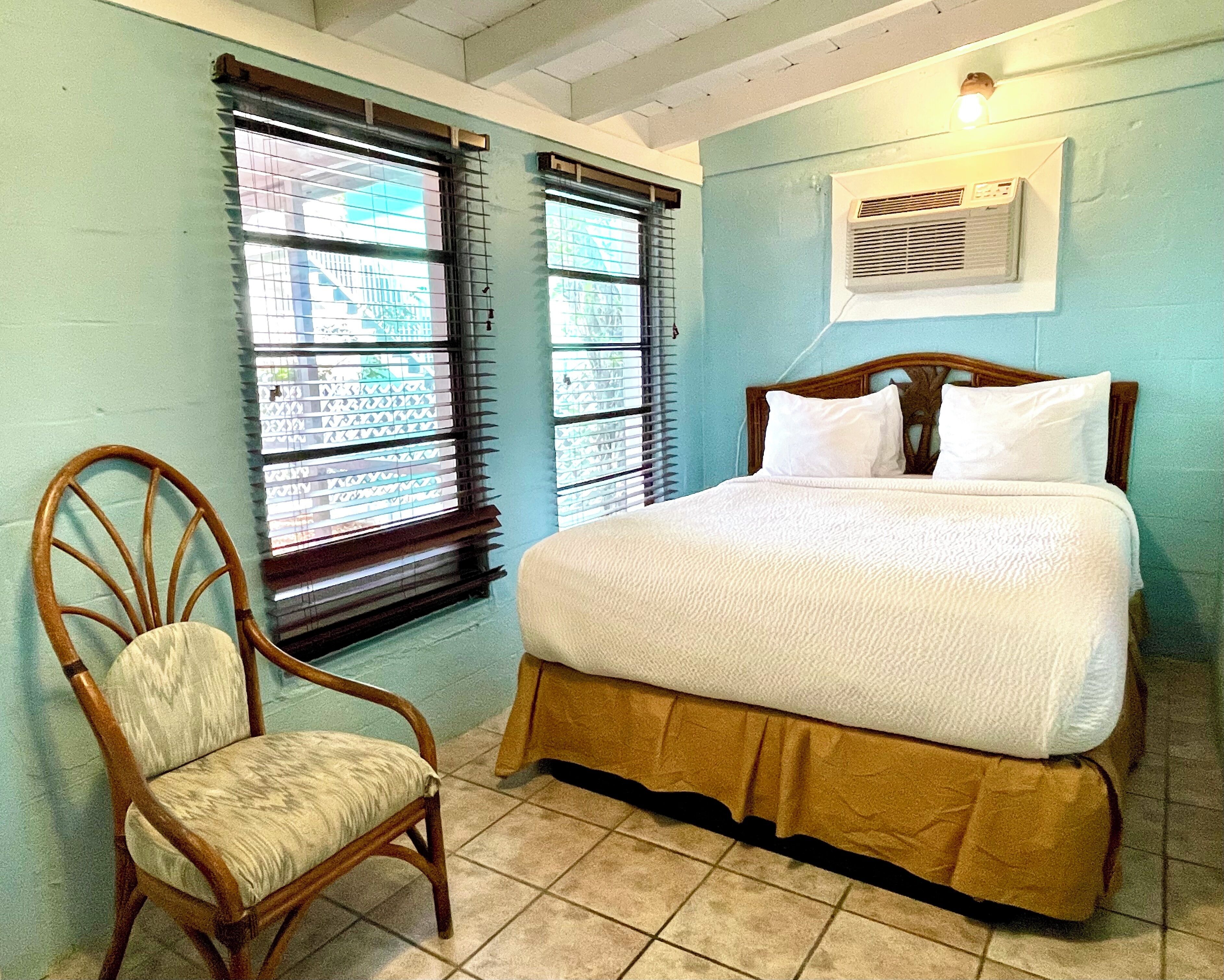 Quarto com cama, uma cadeira e duas janelas com persianas.