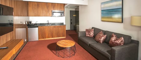 Quarto de hotel moderno com cozinha compacta, sofá cinza com almofadas decorativas, mesa de centro redonda de madeira e uma pintura de uma cena de praia na parede.