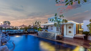 Piscina com espreguiçadeiras e guarda-sóis, um pequeno edifício com parede de pedra, e um céu ao pôr do sol.