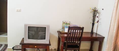 Quarto pequeno com uma mesa de madeira, televisão em um suporte, uma cadeira e uma lixeira.