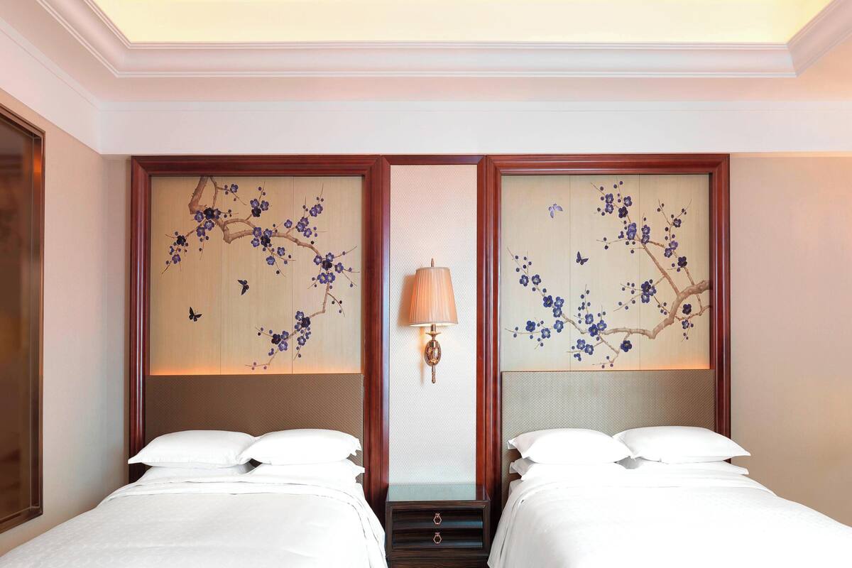 Quarto de hotel com duas camas, uma luminária fixada na parede e arte floral tradicional na cabeceira.