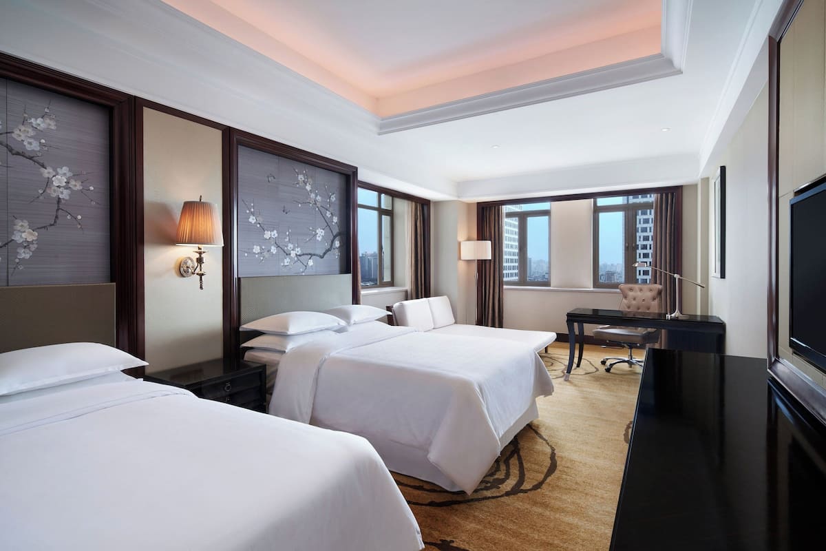 Quarto de hotel com duas camas, uma escrivaninha, televisão e uma janela grande com vista para a cidade.