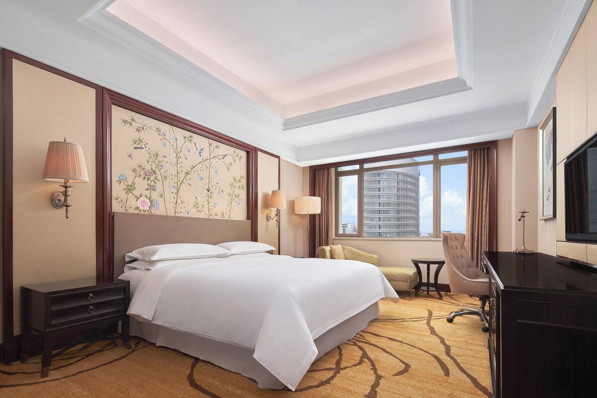 Quarto de hotel com uma cama grande, uma escrivaninha, uma cadeira, uma televisão, um abajur e uma janela com vista para a cidade.