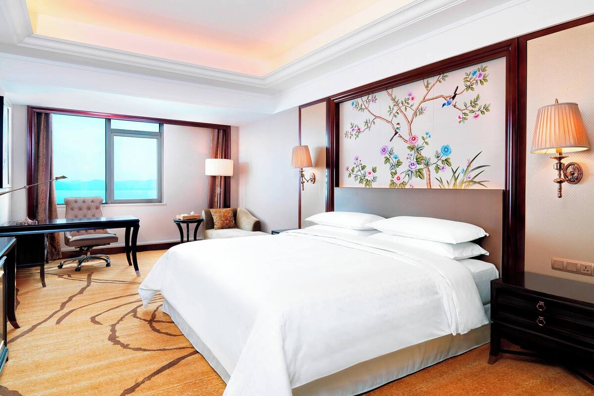 Quarto de hotel com cama grande, escrivaninha, cadeira e vista para o mar.