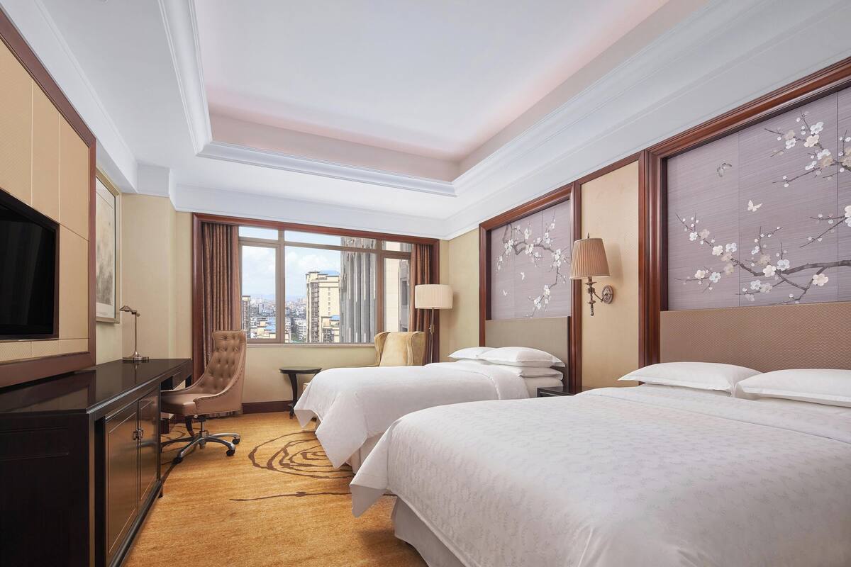 Quarto de hotel com duas camas, uma escrivaninha, uma cadeira, televisão e uma grande janela com vista para a cidade.