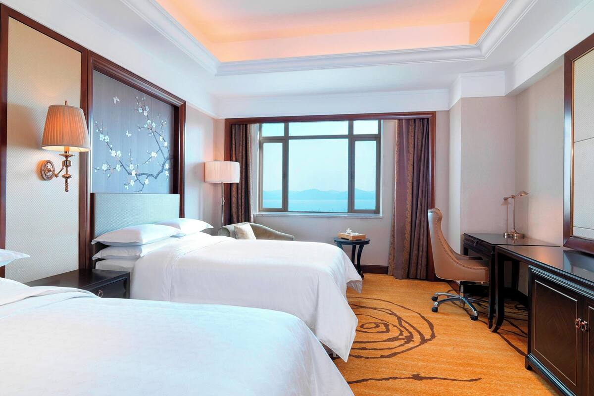 Quarto de hotel com duas camas, uma escrivaninha, uma cadeira, um abajur, um quadro e vista para as montanhas.