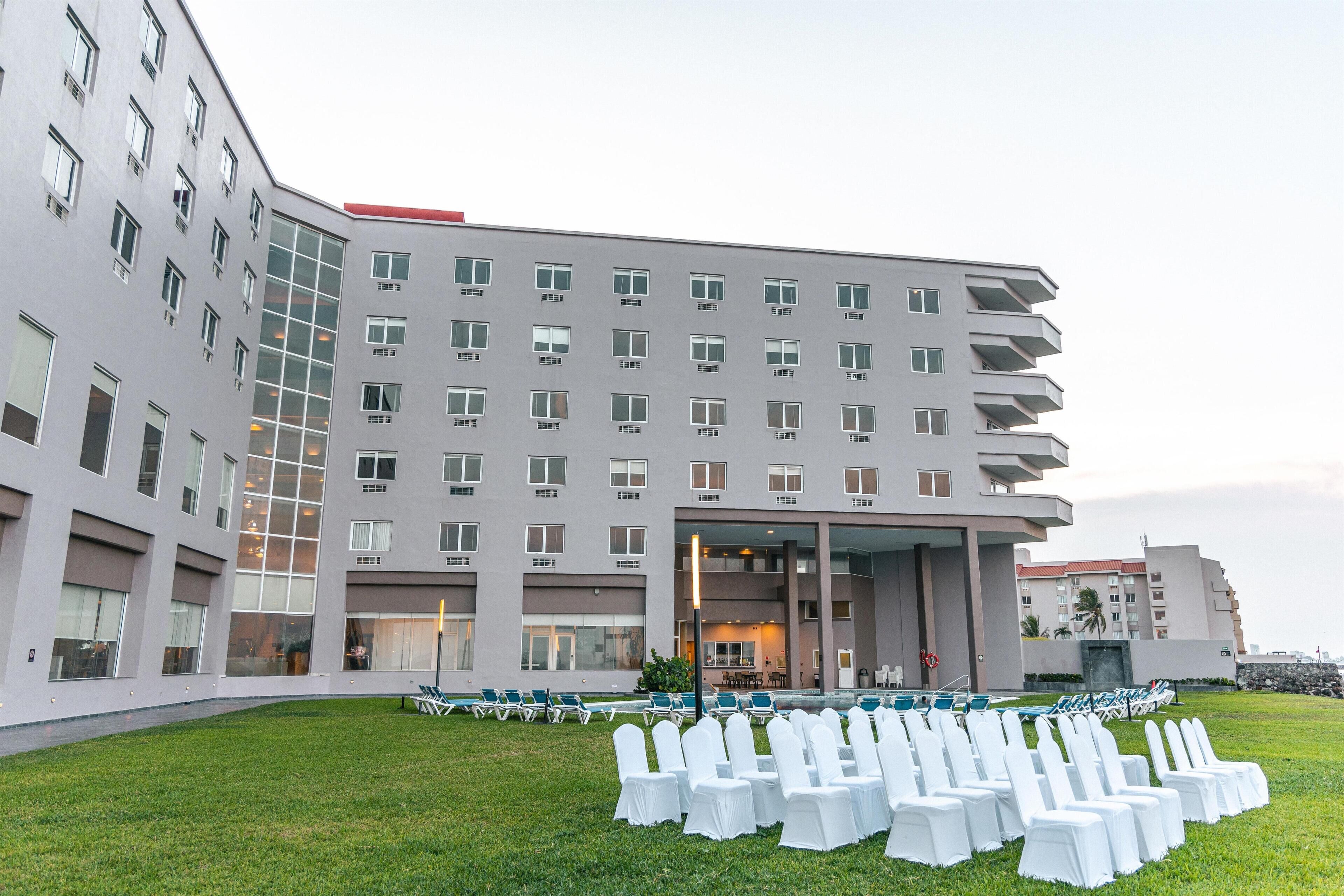 Um hotel moderno com área externa ampla, fileiras de cadeiras brancas e uma piscina.
