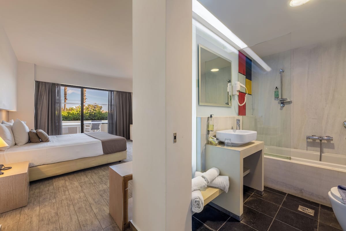 Quarto de hotel moderno com cama, mesa de cabeceira e vista para o exterior.  Ao lado, um banheiro com chuveiro, pia e espelho.