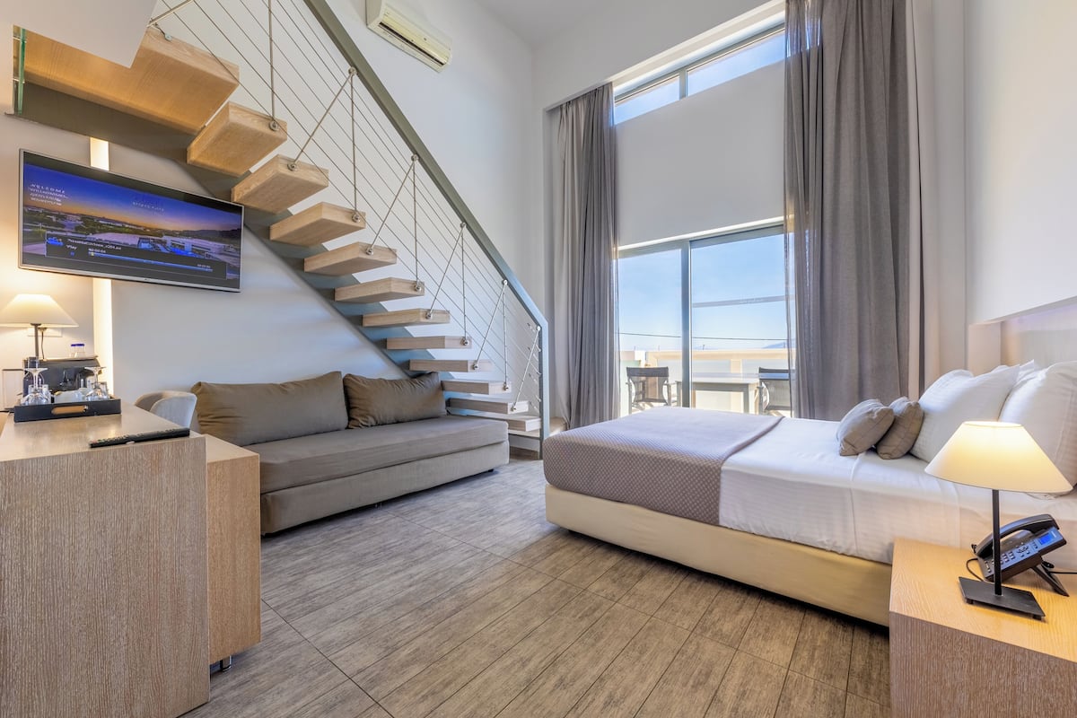 Quarto de hotel moderno com cama embutida, sofá, escrivaninha e escada com tela de TV.