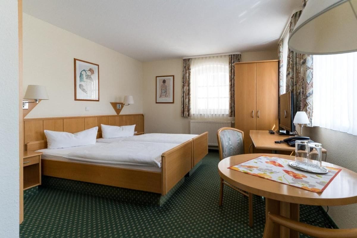 Quarto de hotel com uma cama, escrivaninha, cadeira, mesa e janela com cortinas.