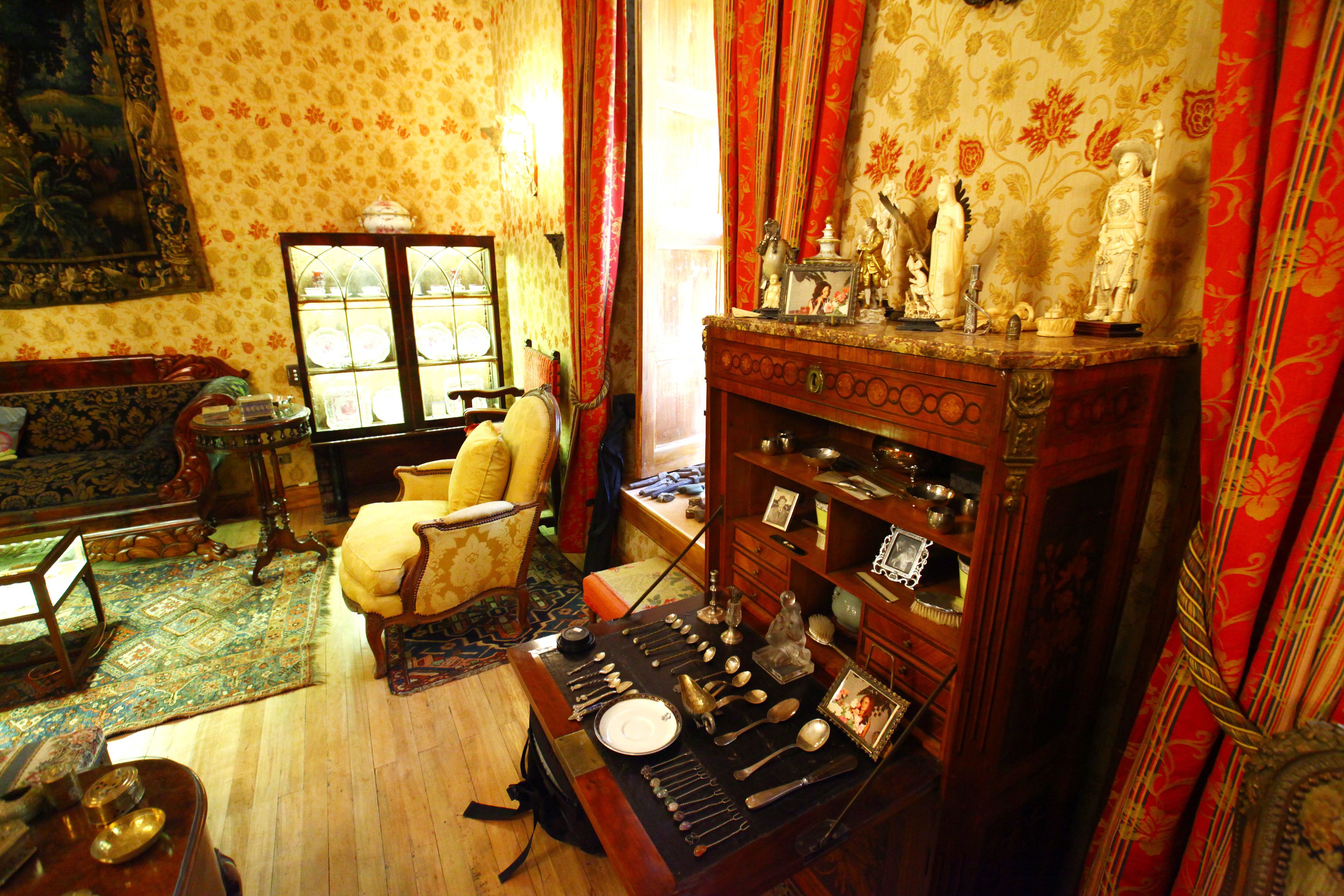 Quarto com armário de madeira exibindo diversos objetos, uma poltrona amarela, tapete estampado e janela com cortinas.