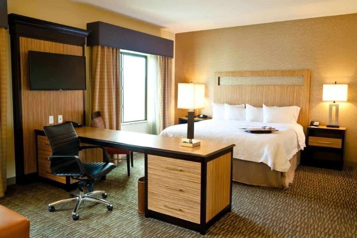 Quarto de hotel com cama, escrivaninha, cadeira, televisão, luminárias e uma janela.