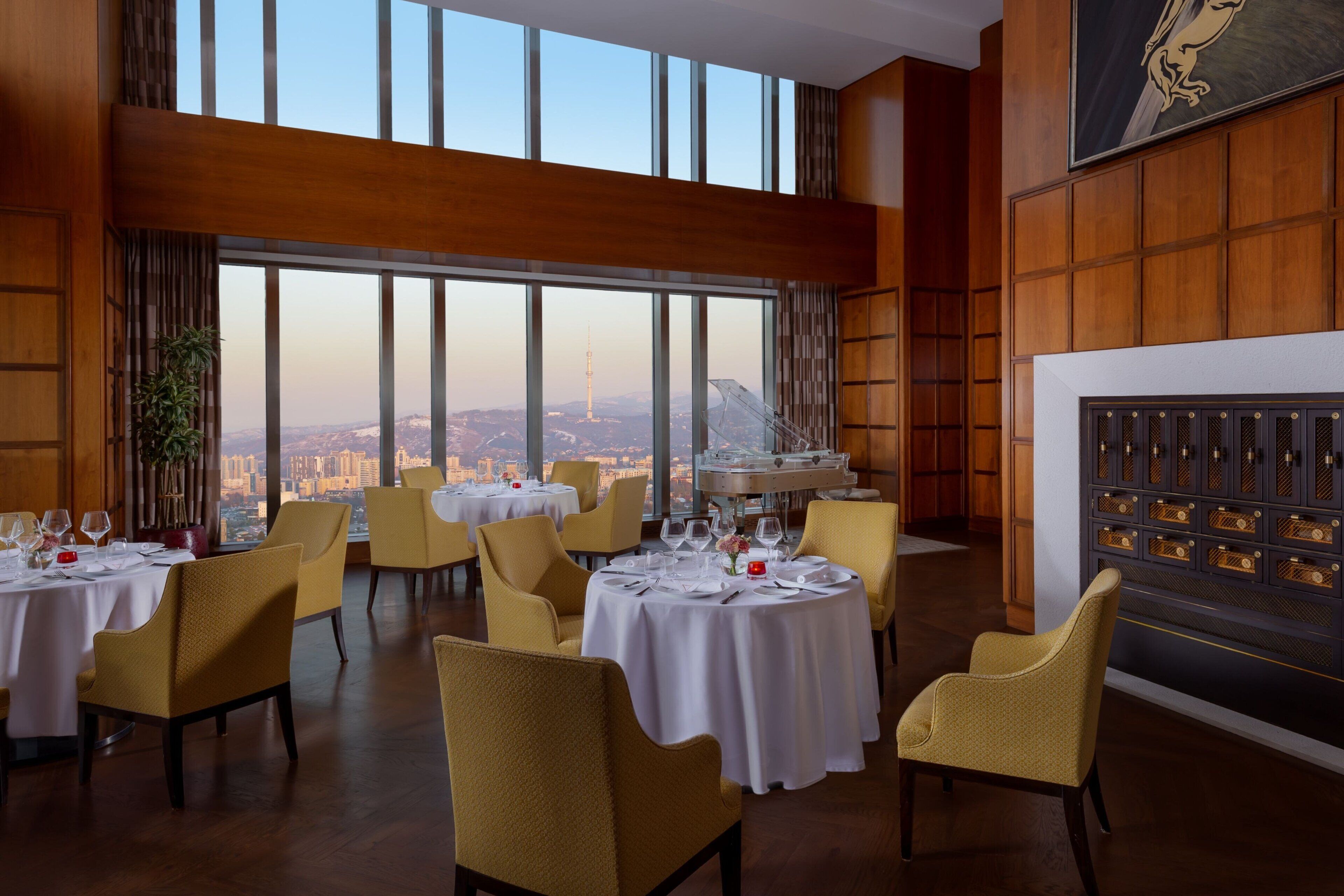Restaurante com mesas redondas preparadas para refeições, grandes janelas com vista para a cidade e uma adega na parede.