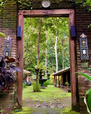 Um arco de madeira rústico que dá acesso a uma área externa coberta, com jardim e uma fonte.
