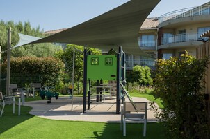 Um parquinho com uma estrutura verde para escalar, balanços e uma área sombreada com toldo.