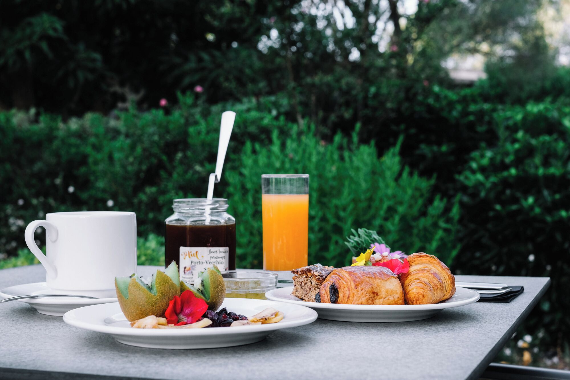Uma mesa com uma caneca branca, um copo de sumo de laranja, um frasco de compota, um prato com croissants e fruta, e outro prato com frutos secos e uma flor.