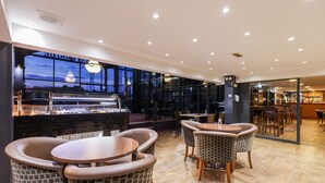 Um restaurante moderno com mesas redondas e cadeiras estofadas, um balcão com vitrine e vista para a paisagem urbana ao entardecer.