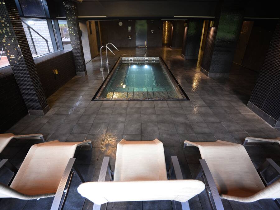 Uma área interna de piscina moderna com barreira de vidro, duas espreguiçadeiras e piso revestido de azulejos.