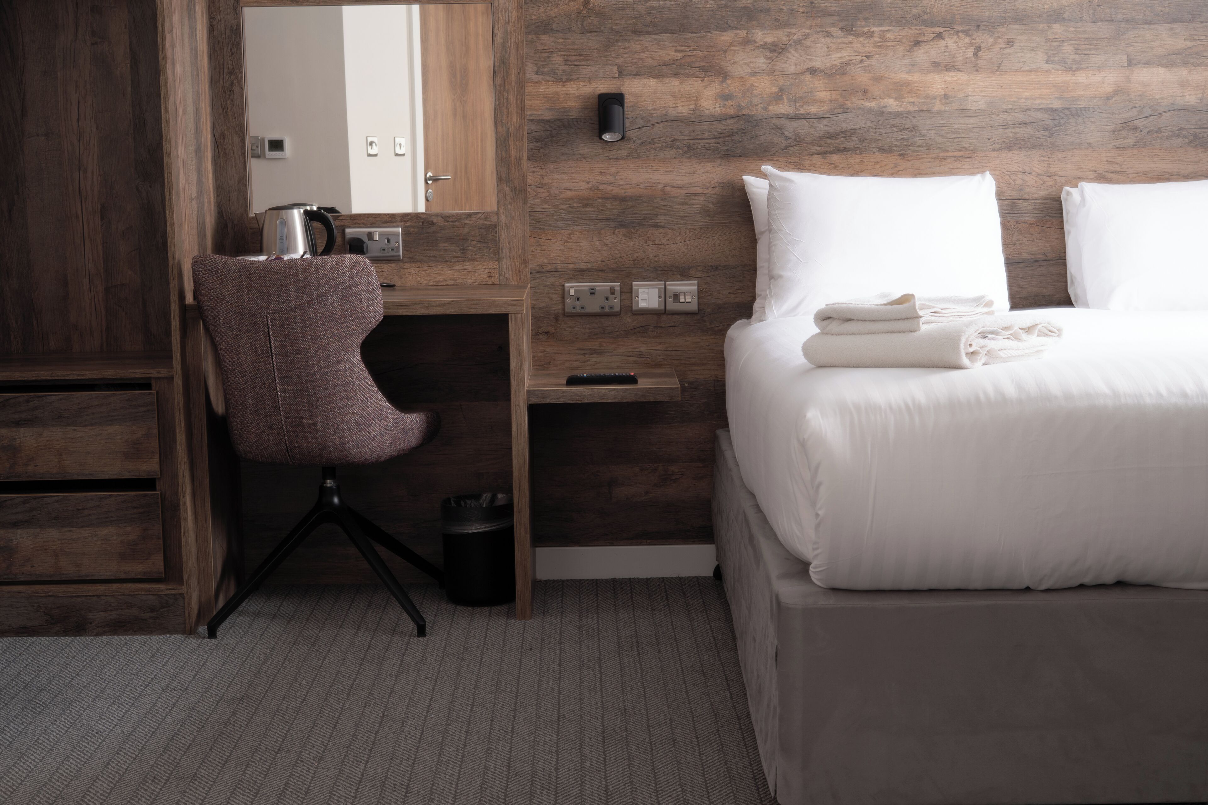 Quarto com cabeceira de madeira, cama com roupa de cama branca, escrivaninha com cadeira e pequena mesa de cabeceira com chaleira e controle remoto.