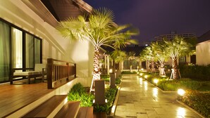 Área externa bem iluminada, com bancos de madeira e palmeiras, que dá acesso a um edifício.