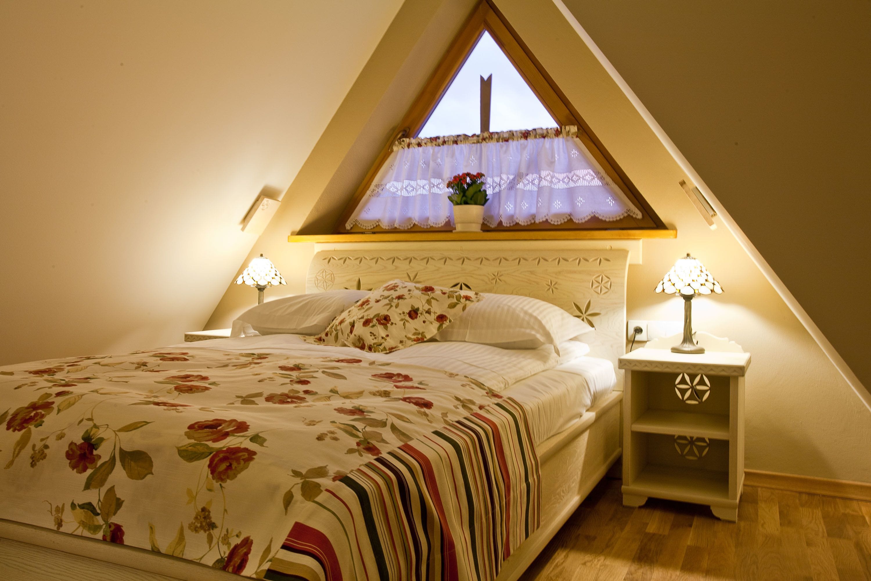 Quarto com teto inclinado, cama com roupa de cama florida, duas luminárias de cabeceira e mesa de cabeceira com um objeto decorativo.