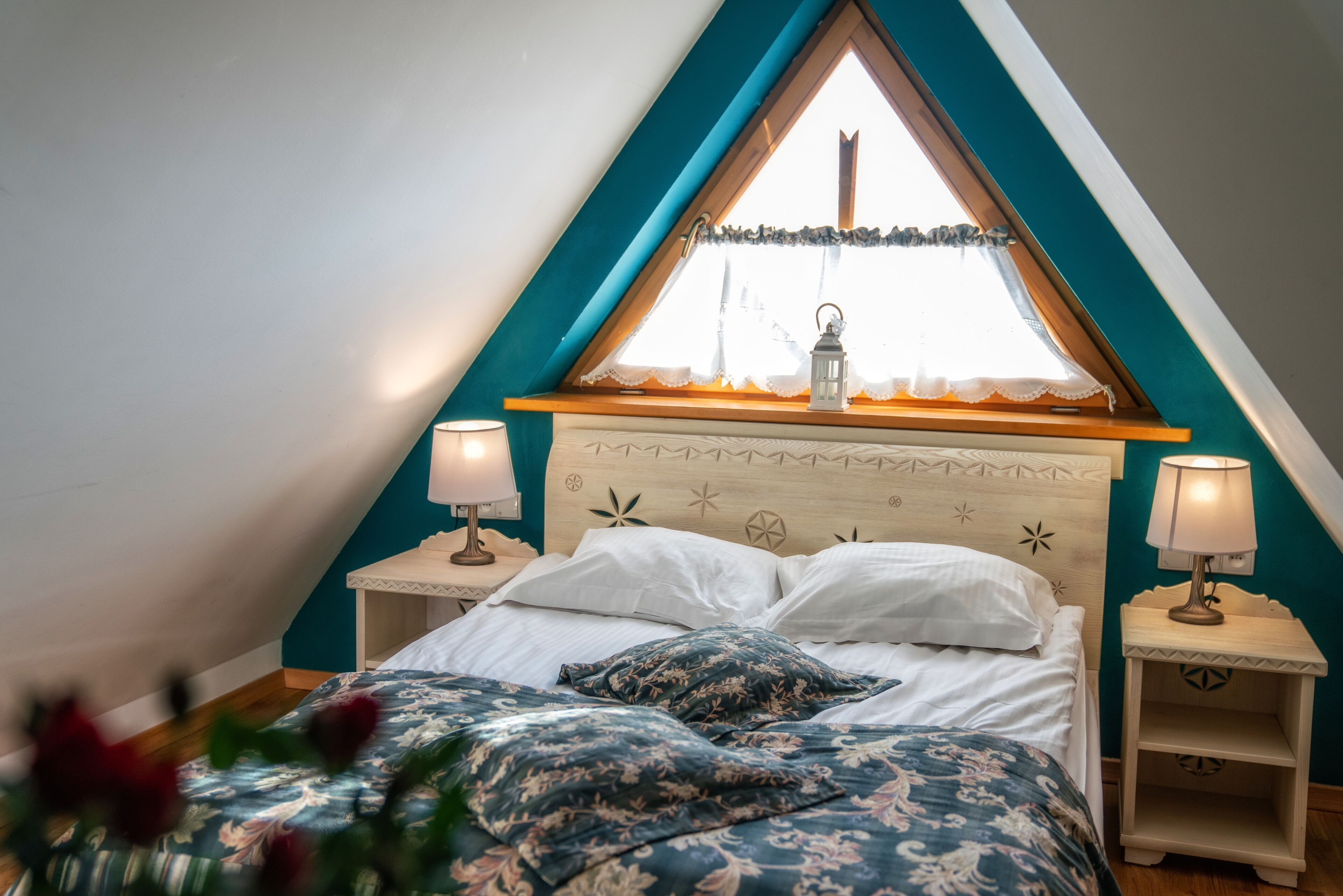 Quarto com teto inclinado, cama com roupa branca, duas mesas de cabeceira com abajures e uma janela com lanterneiro.