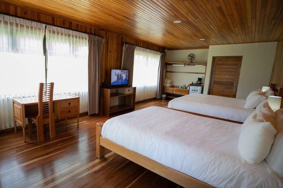 Quarto de hotel com piso de madeira, uma escrivaninha com cadeira, televisão, duas camas e um guarda-roupa.