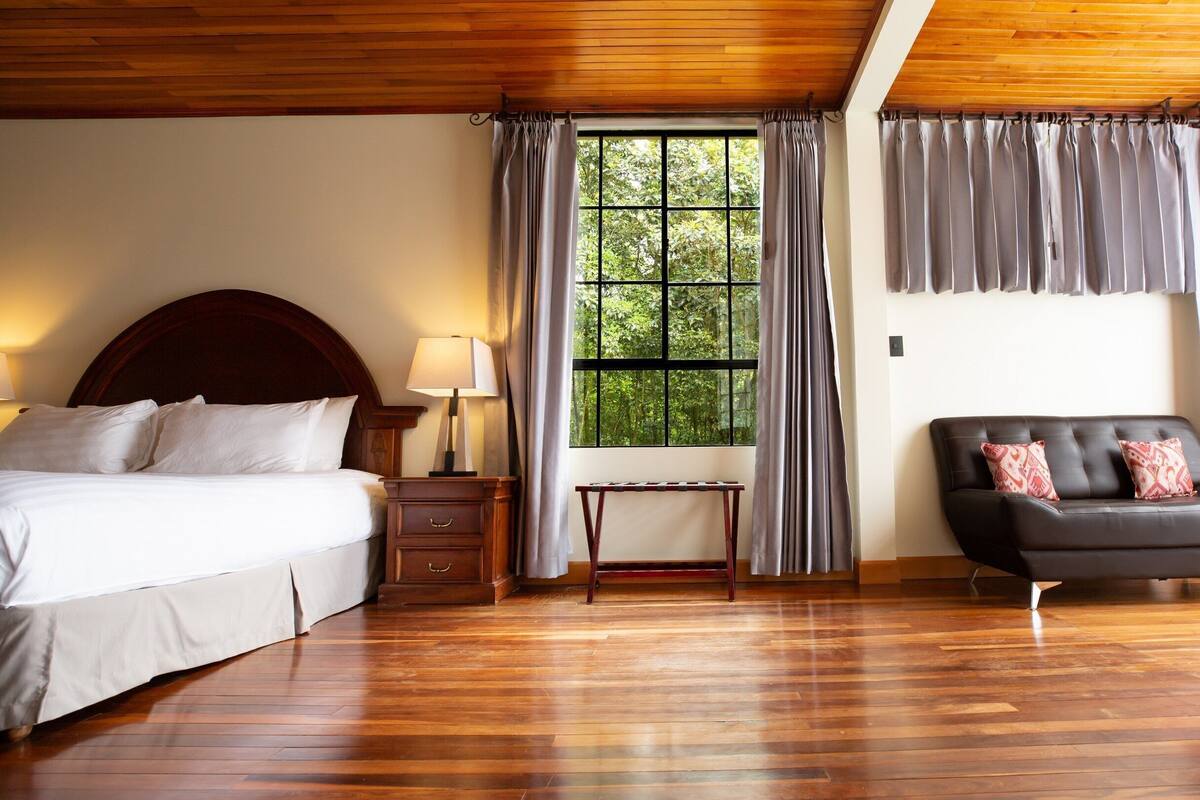 Quarto de hotel com teto de madeira, uma janela grande com cortinas, uma cama, uma mesinha de cabeceira, uma mesa e um sofá.