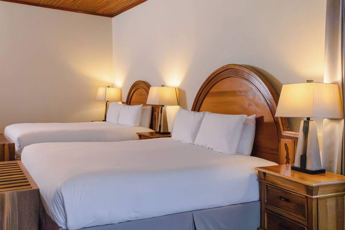 Quarto de hotel com duas camas, cabeceiras de madeira, mesas de cabeceira com abajures e paleta de cores neutras.