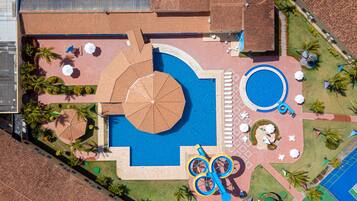Vista aérea de um resort com piscina central, diversos guarda-sóis, área de recreação infantil e quadra de tênis.