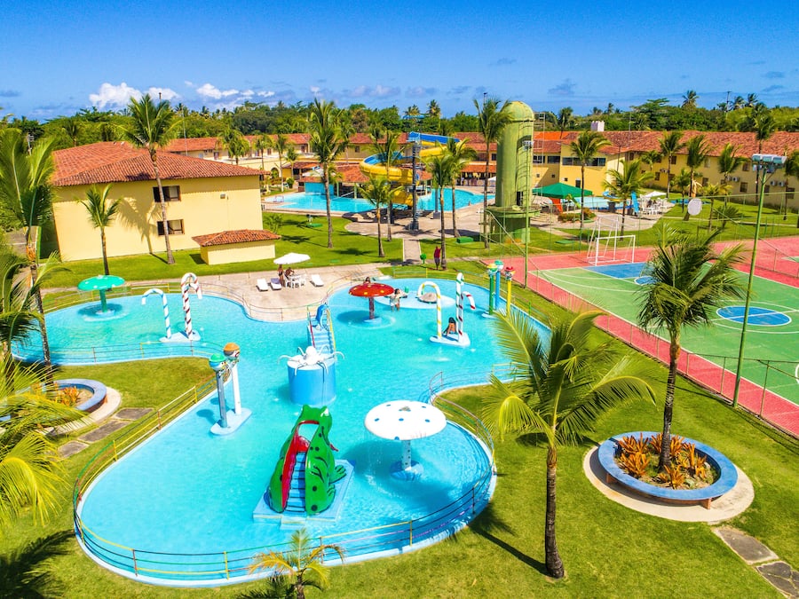 Um parque aquático com várias piscinas, toboáguas e uma quadra de basquete, tudo cercado por palmeiras e edifícios.