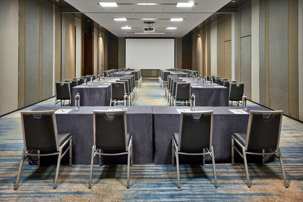 Sala de conferências com mesas e cadeiras dispostas em fileiras, uma tela de projetor e piso coberto por carpete.