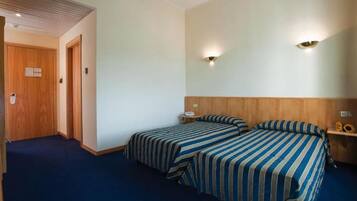 Quarto de hotel com duas camas, cabeceira de madeira, mesa de cabeceira e luminária fixada na parede.
