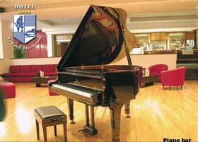 Um piano de cauda em um saguão de hotel, com um sofá vermelho e um bar ao fundo.
