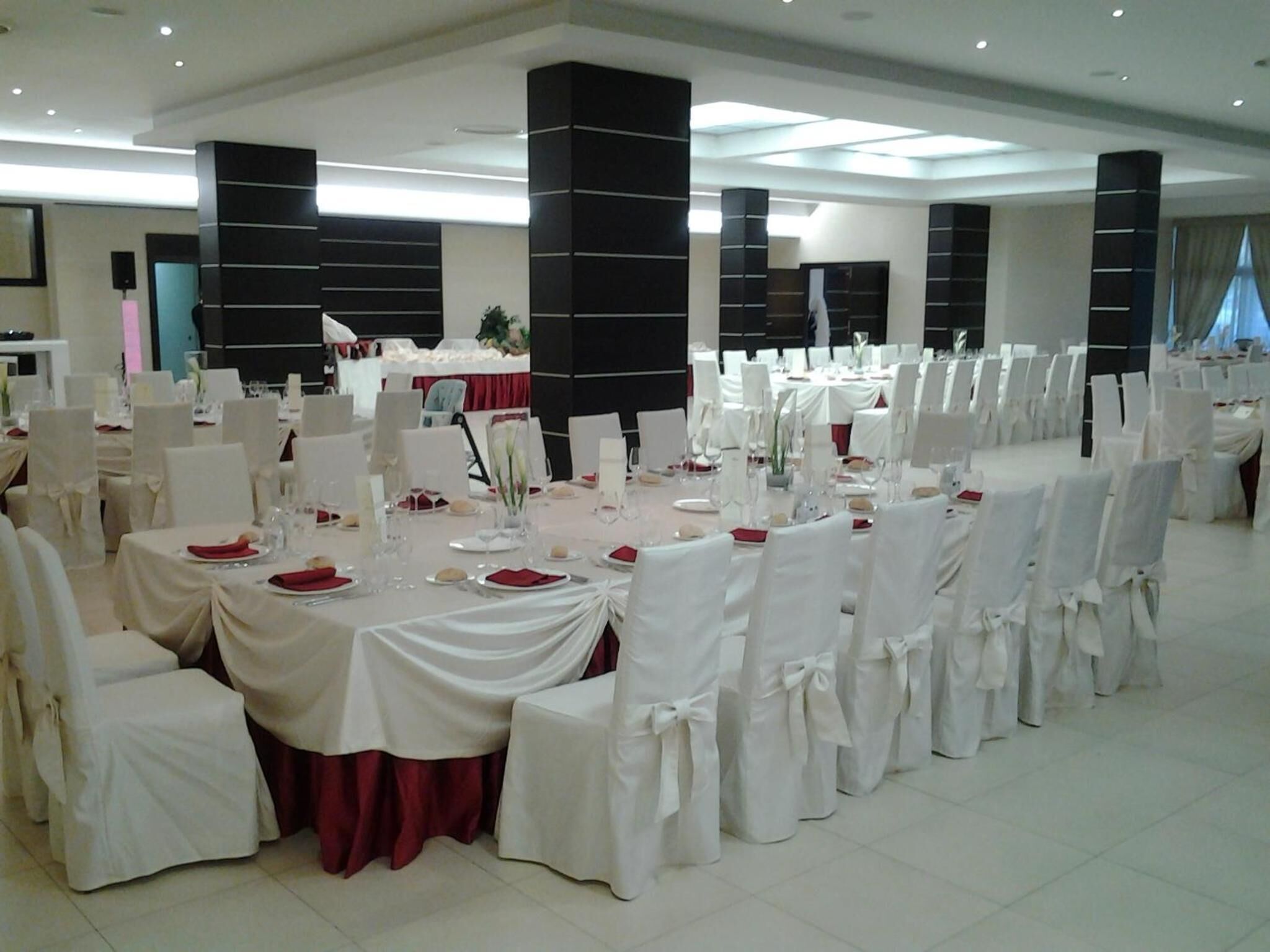 Uma sala de banquetes com mesas redondas dispostas para um evento formal, com toalhas brancas, guardanapos vermelhos e cadeiras brancas.