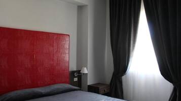 Quarto de hotel com cabeceira vermelha, cama com roupa de cama cinzenta, mesa de cabeceira com abajur e janela com cortinas escuras.